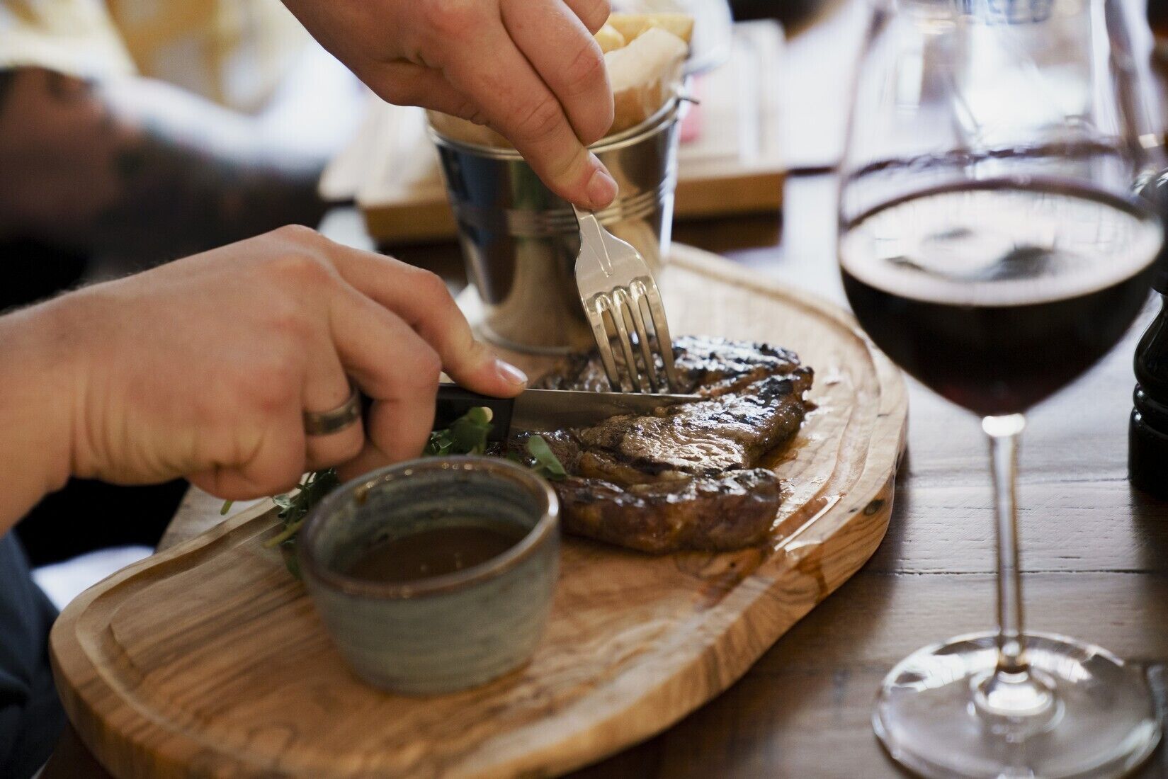 Uma pessoa cortando bife em tábua de madeira, garfo e faca, uma pequena tigela de molho e um copo de vinho tinto.