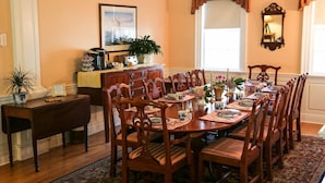 Uma sala de jantar com uma mesa redonda posta para seis pessoas, rodeada por cadeiras de madeira. Um aparador de madeira está encostado à parede, e há um vaso de planta sobre uma mesinha lateral.