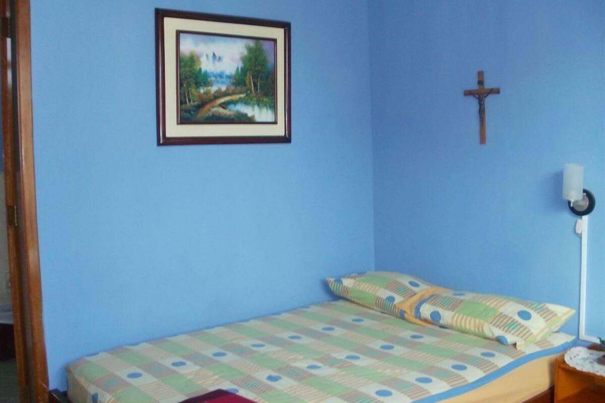 Quarto com cama, um quadro emoldurado, uma cruz e um abajur.