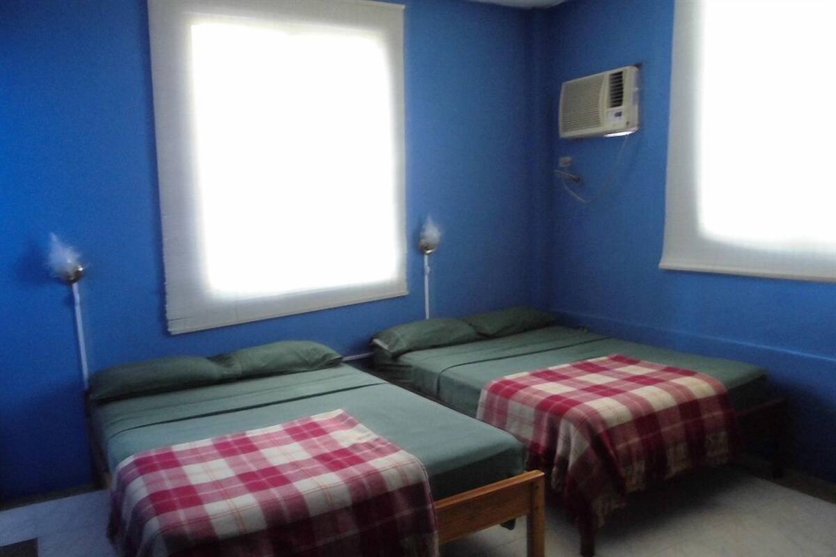 Quarto com duas camas, cada uma com colcha verde e manta xadrez vermelha e branca. Há uma janela com persianas brancas, um ar condicionado na parede e uma pequena mesa com abajur.