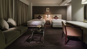 Quarto de hotel com duas camas, uma escrivaninha, uma cadeira, uma mesa de centro e uma janela grande com cortinas.