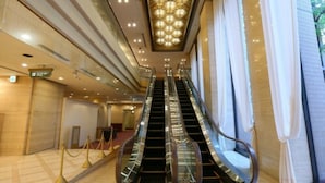 Interior moderno de hotel com um escalator central que leva ao segundo andar, rodeado por grades de vidro e um lustre decorativo.