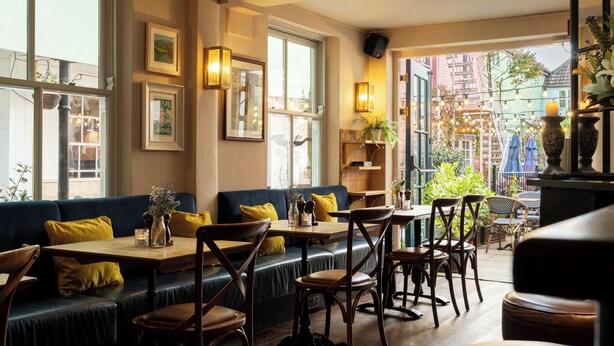Interior acolhedor de restaurante com sofá azul, mesas e cadeiras de madeira, e amplas janelas com vista para a cidade.