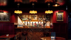 Uma área de bar com piso de madeira em padrão herringbone, banquetas e diversos frascos e copos nas prateleiras.