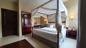 Quarto de hotel com cama de dossel, escrivaninha de madeira, mesa de cabeceira, abajur e porta.