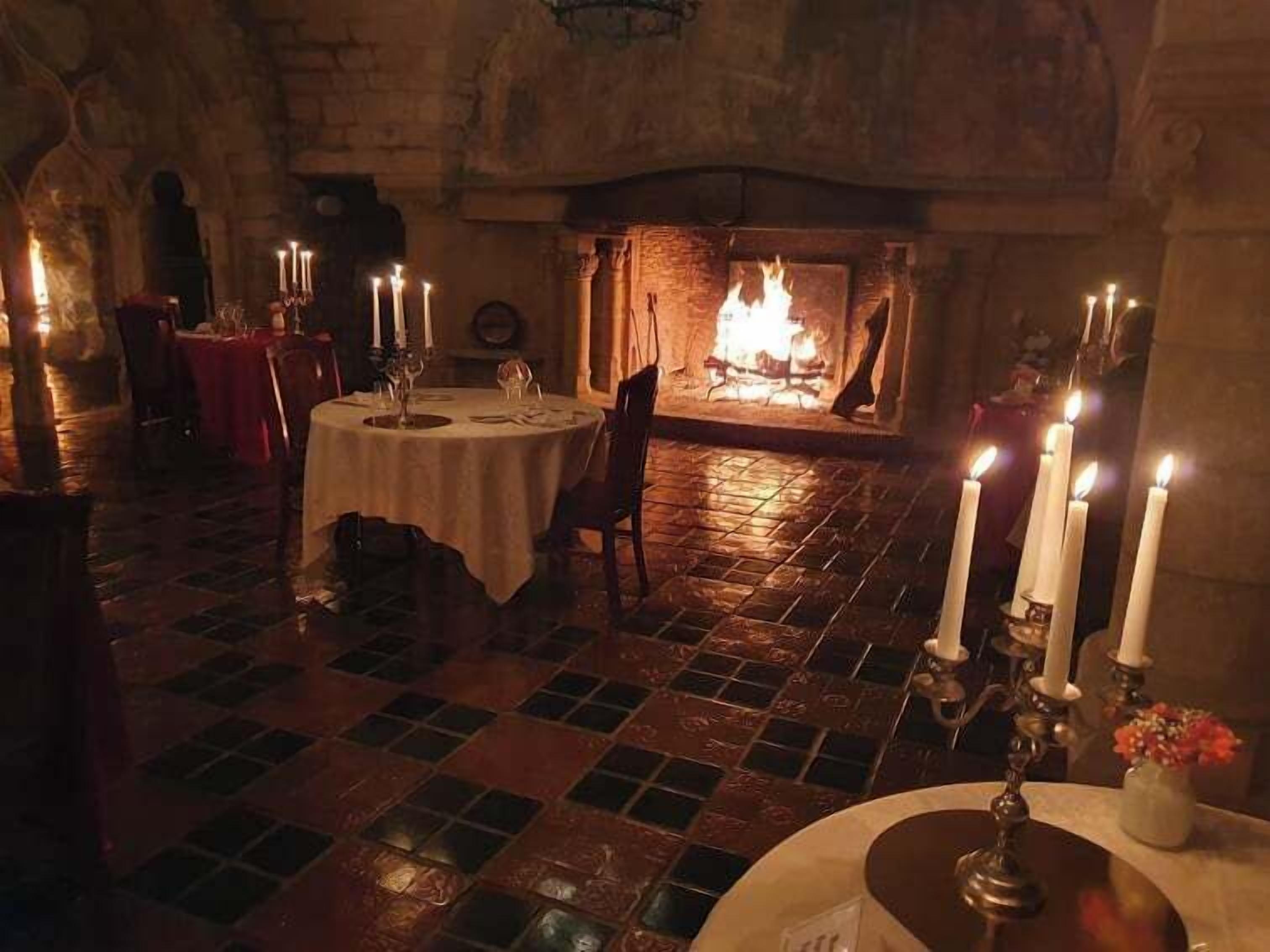 Um quarto com pouca luz, lareira acesa, uma mesa redonda posta para refeição e várias velas acesas.