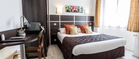 Quarto de hotel moderno com uma cama grande, uma escrivaninha com cadeira, uma mesinha e uma mesa de cabeceira. Há luminárias fixadas na parede e uma janela com cortinas.