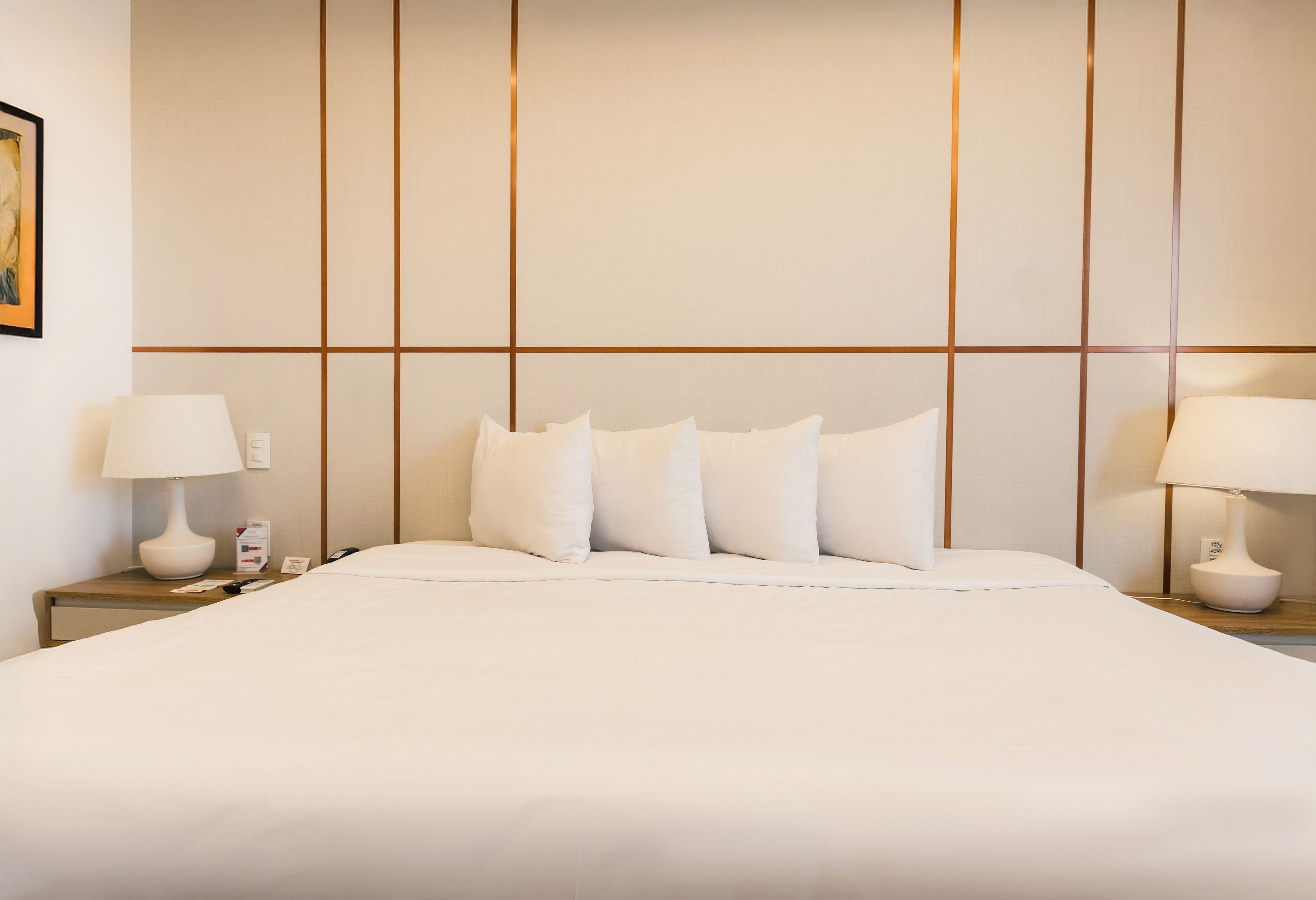 Quarto de hotel com uma cama grande, duas mesas de cabeceira com luminárias e uma parede lisa com linhas verticais.
