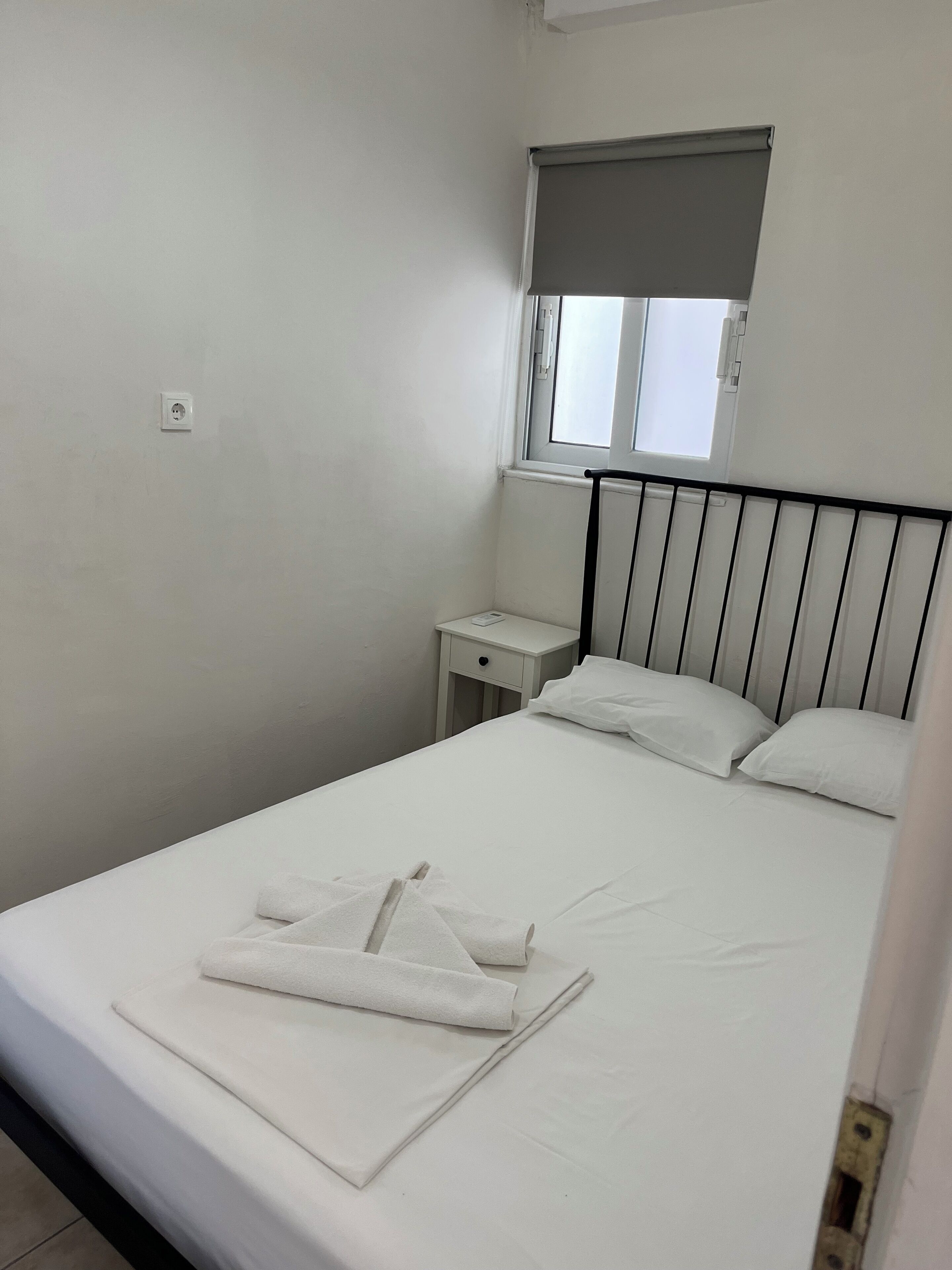 Uma cama de solteiro com colchão e almofadas brancos, uma mesinha de cabeceira com um telefone, uma janela com persiana e uma tomada na parede.