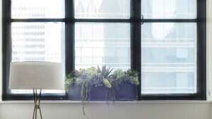 Quarto moderno com poltrona azul escura, luminária de chão e janela com vista para edifícios e um vaso com plantas.