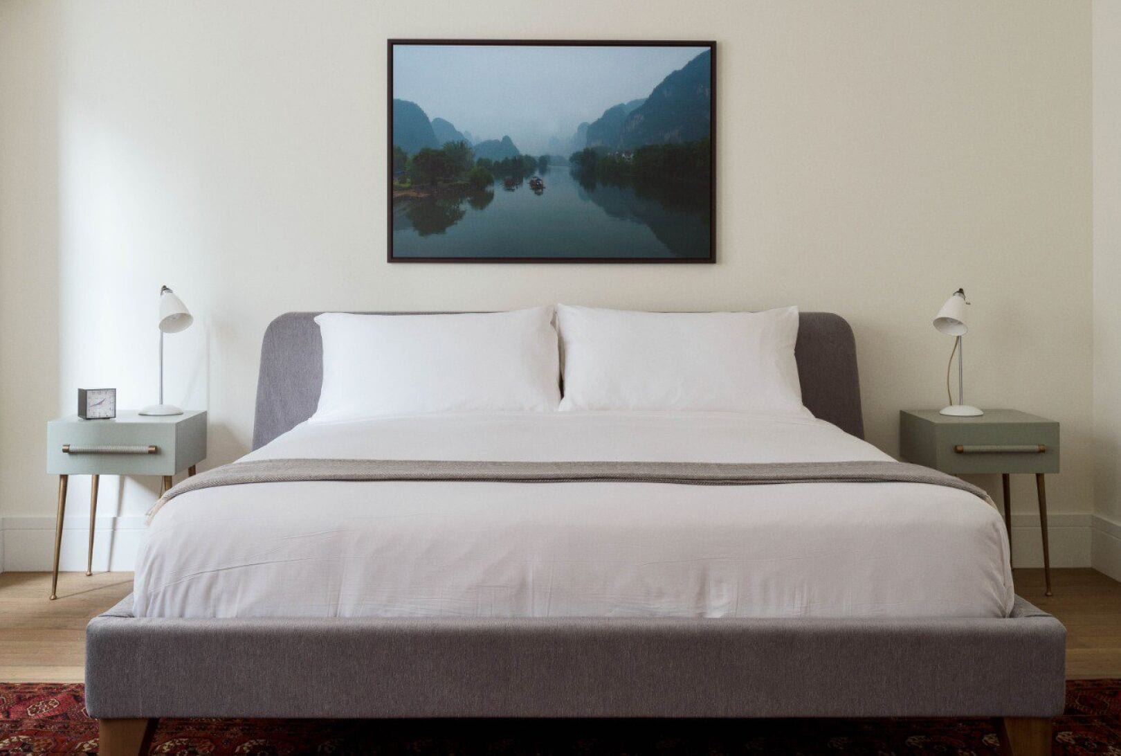Uma cama grande com cabeceira cinzenta e roupa de cama branca, acompanhada por duas mesas de cabeceira com abajures, em um quarto com uma pintura paisagística emoldurada acima.