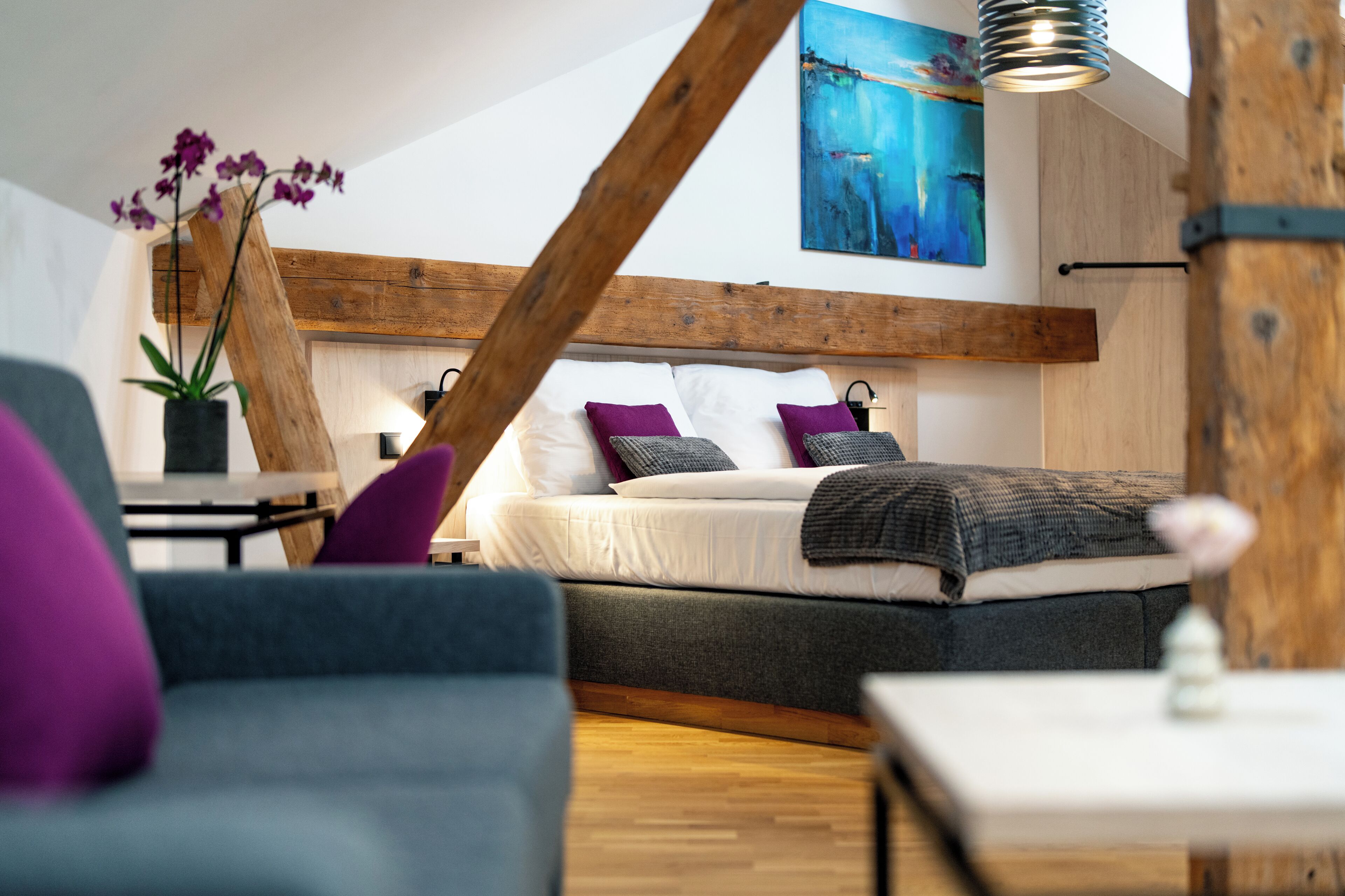 Quarto moderno com teto de madeira, cama com roupa de cama branca e almofadas roxas, uma pequena mesa com abajur e um quadro na parede.