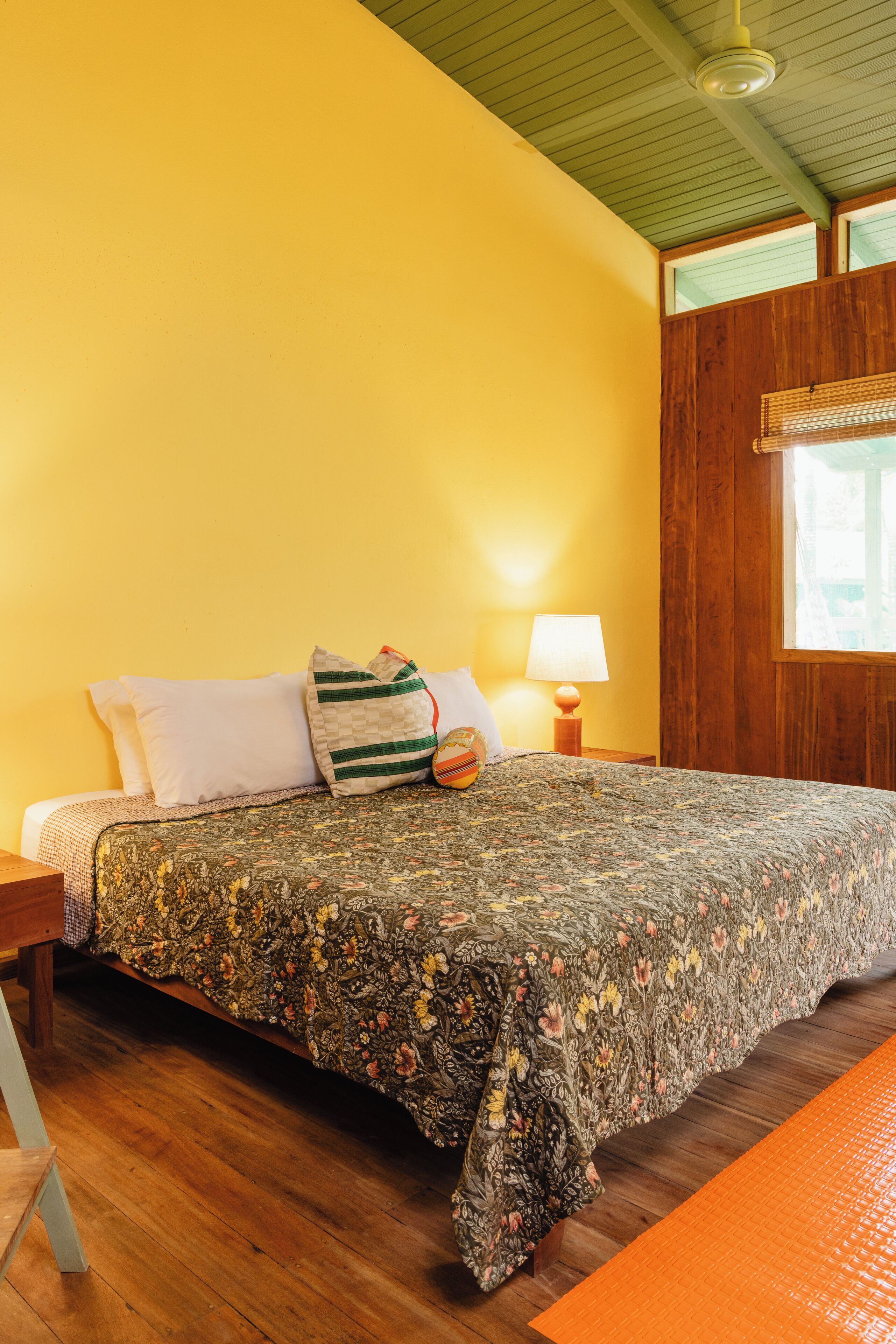 Quarto com uma cama, piso de madeira, parede amarela, janela e uma luminária.