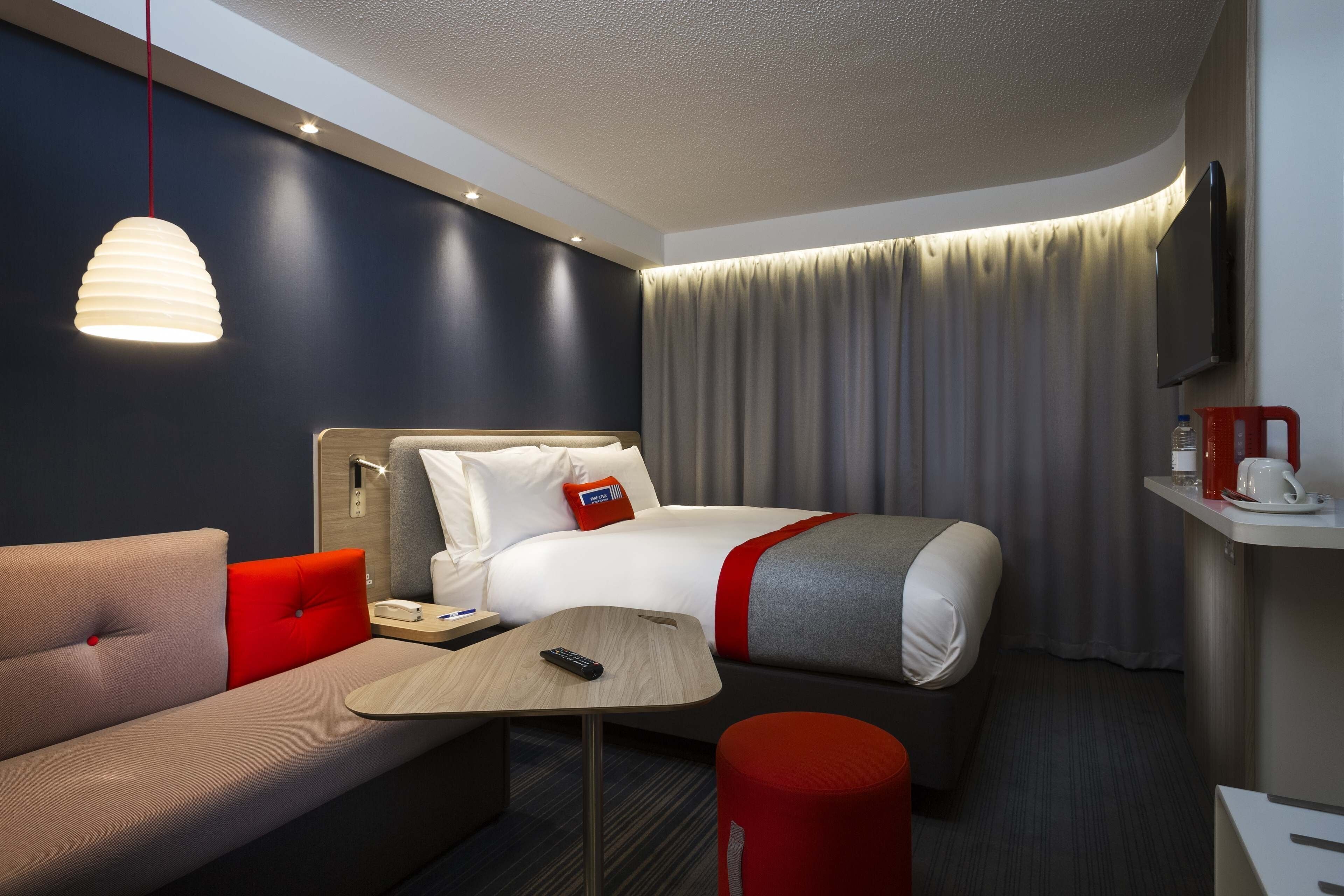 Quarto de hotel com cama, sofá, mesinha, banquinho vermelho, televisão e cortina.