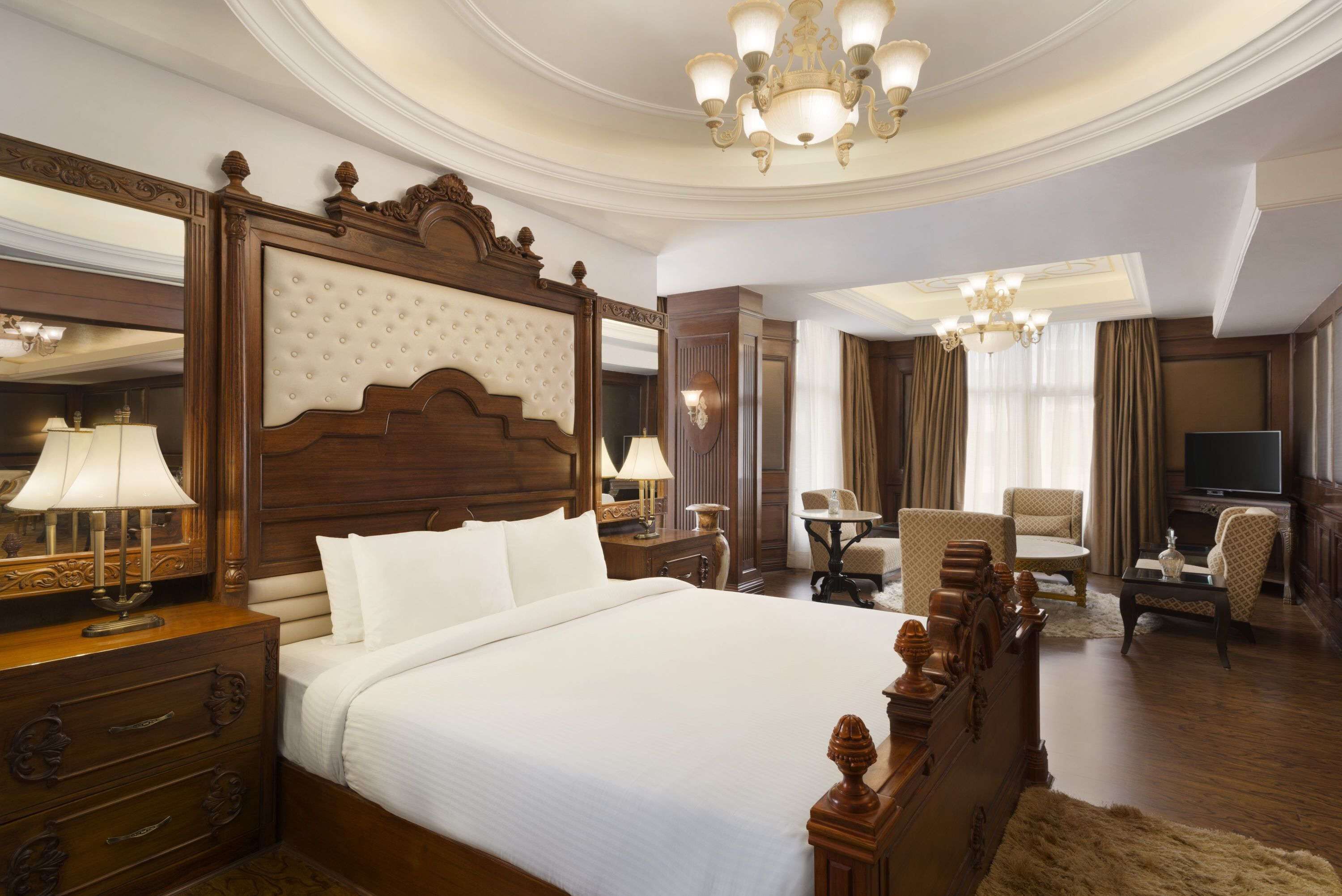 Quarto de hotel com uma cama grande, cabeceira de madeira, duas mesas de cabeceira com luminárias, uma cadeira, uma televisão e um candelabro.