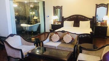 Quarto de hotel com uma cama grande, duas poltronas, um sofá, um banheiro com paredes de vidro e piso de madeira.