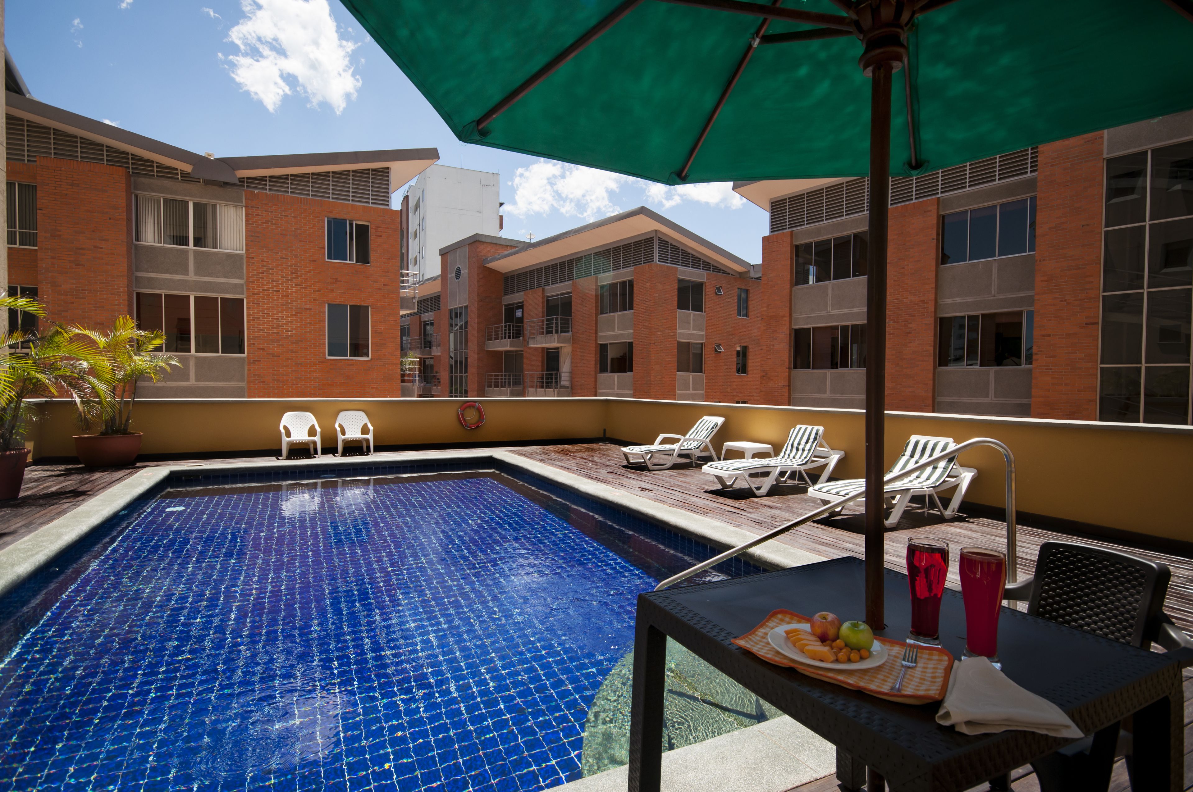 Área de piscina no terraço, com piscina azulejada em azul, espreguiçadeiras e uma mesa com tábua de frutas e bebidas.