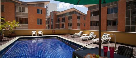 Área de piscina no terraço, com piscina azulejada em azul, espreguiçadeiras e uma mesa com tábua de frutas e bebidas.