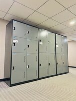 Uma fileira de armários metálicos cinzas com etiquetas numeradas, teto com iluminação embutida e piso coberto de carpete.