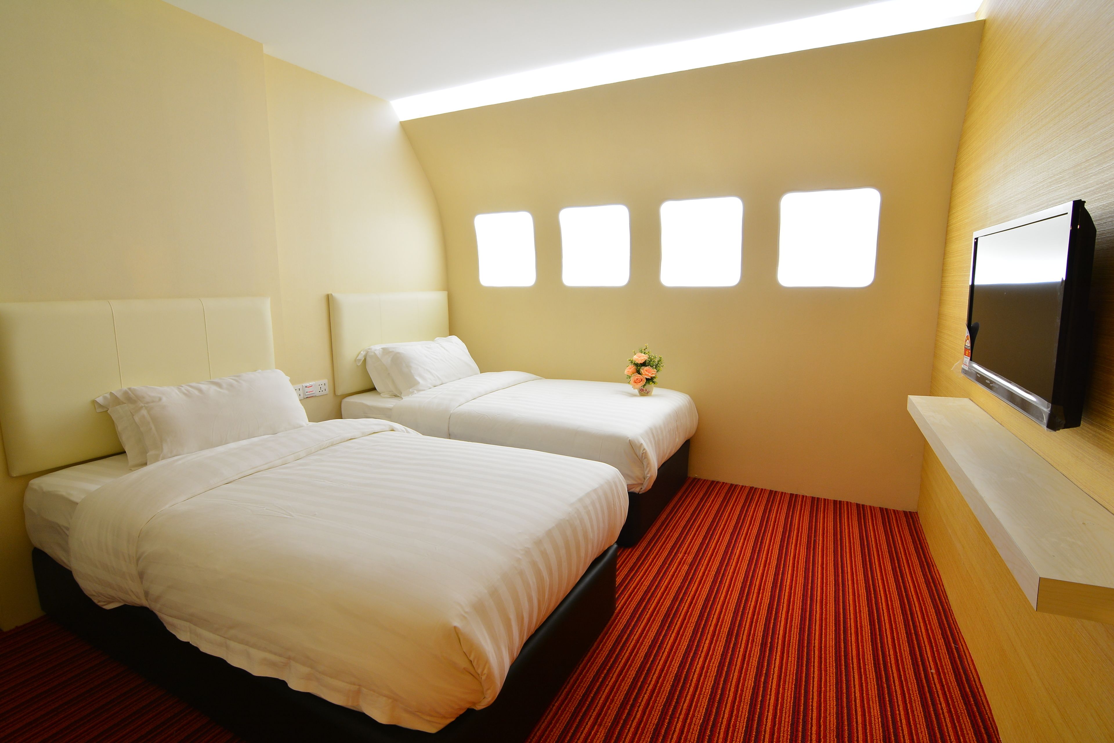 Quarto de hotel com duas camas, um tapete vermelho, uma TV e uma janela com três luzes quadradas.