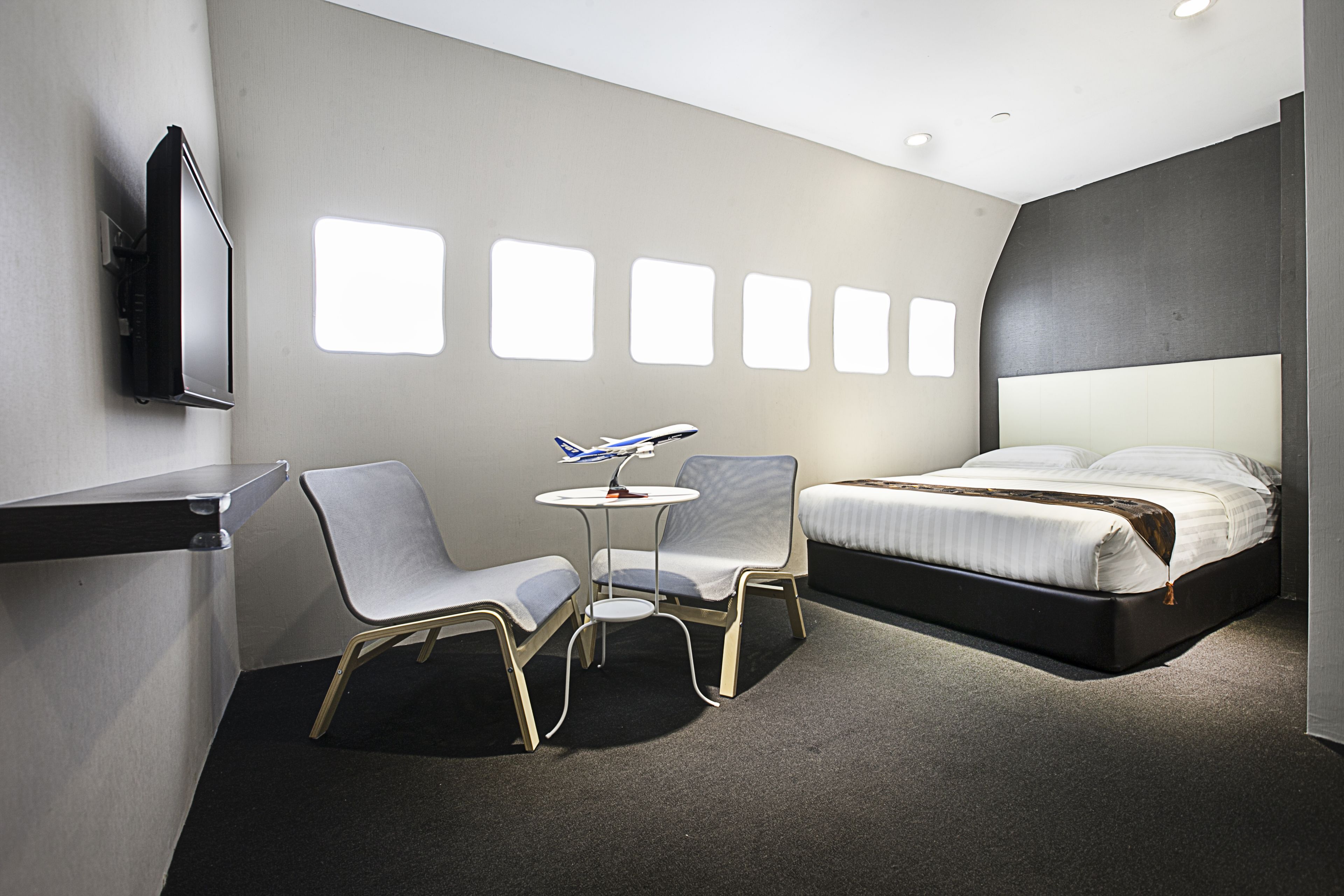 Quarto de hotel moderno com uma cama, duas cadeiras, uma mesinha e uma TV fixada na parede.