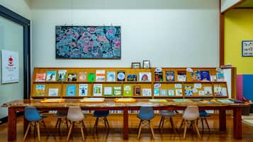 Uma biblioteca com uma longa mesa de madeira e várias cadeiras. Estantes com livros e uma obra de arte colorida são visíveis.