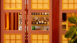Visão através de uma porta de madeira com painéis de vidro, mostrando prateleiras repletas de diversas garrafas e um relógio.