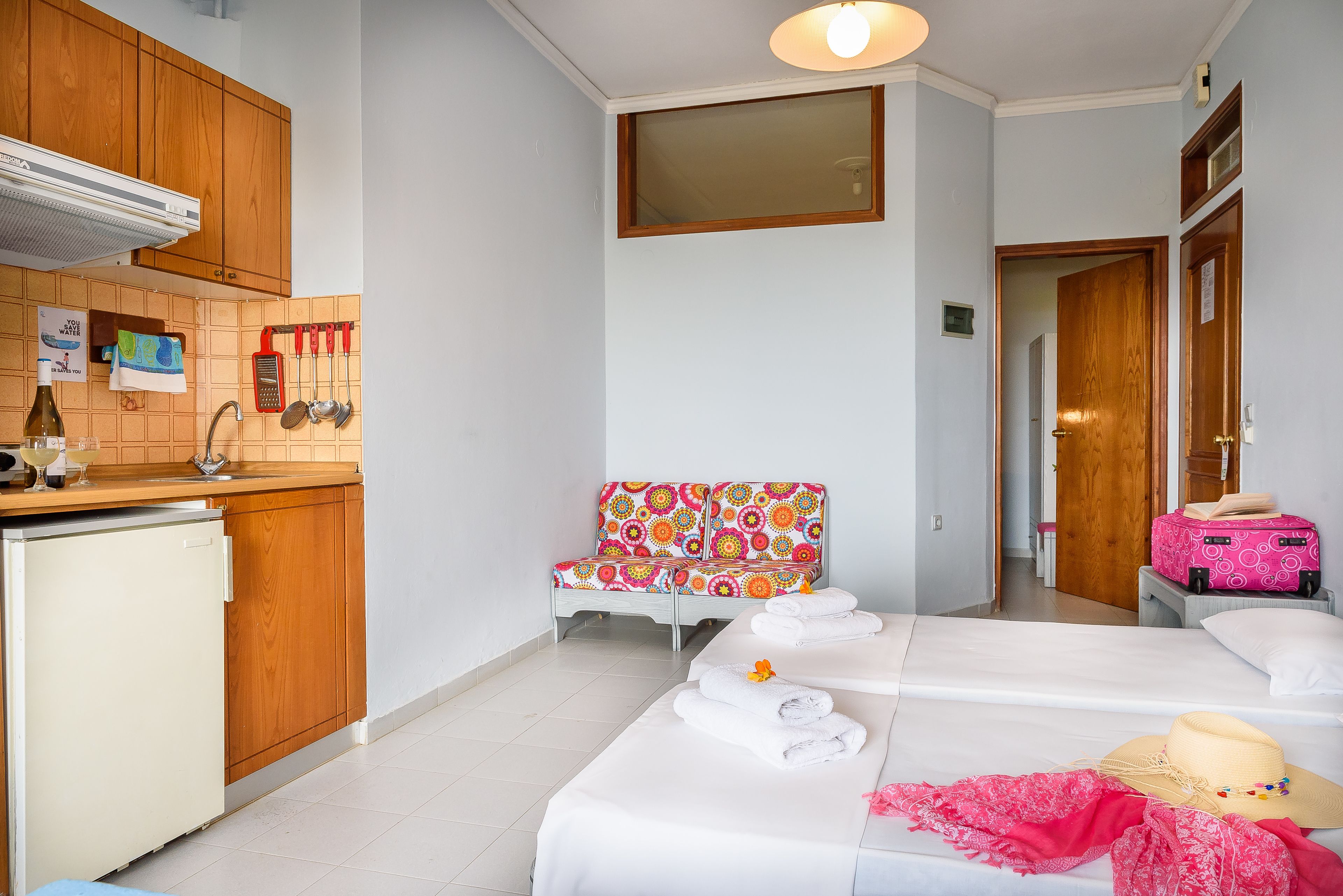 Quarto de hotel pequeno com cozinha compacta, uma cama, uma cadeira e uma mala.