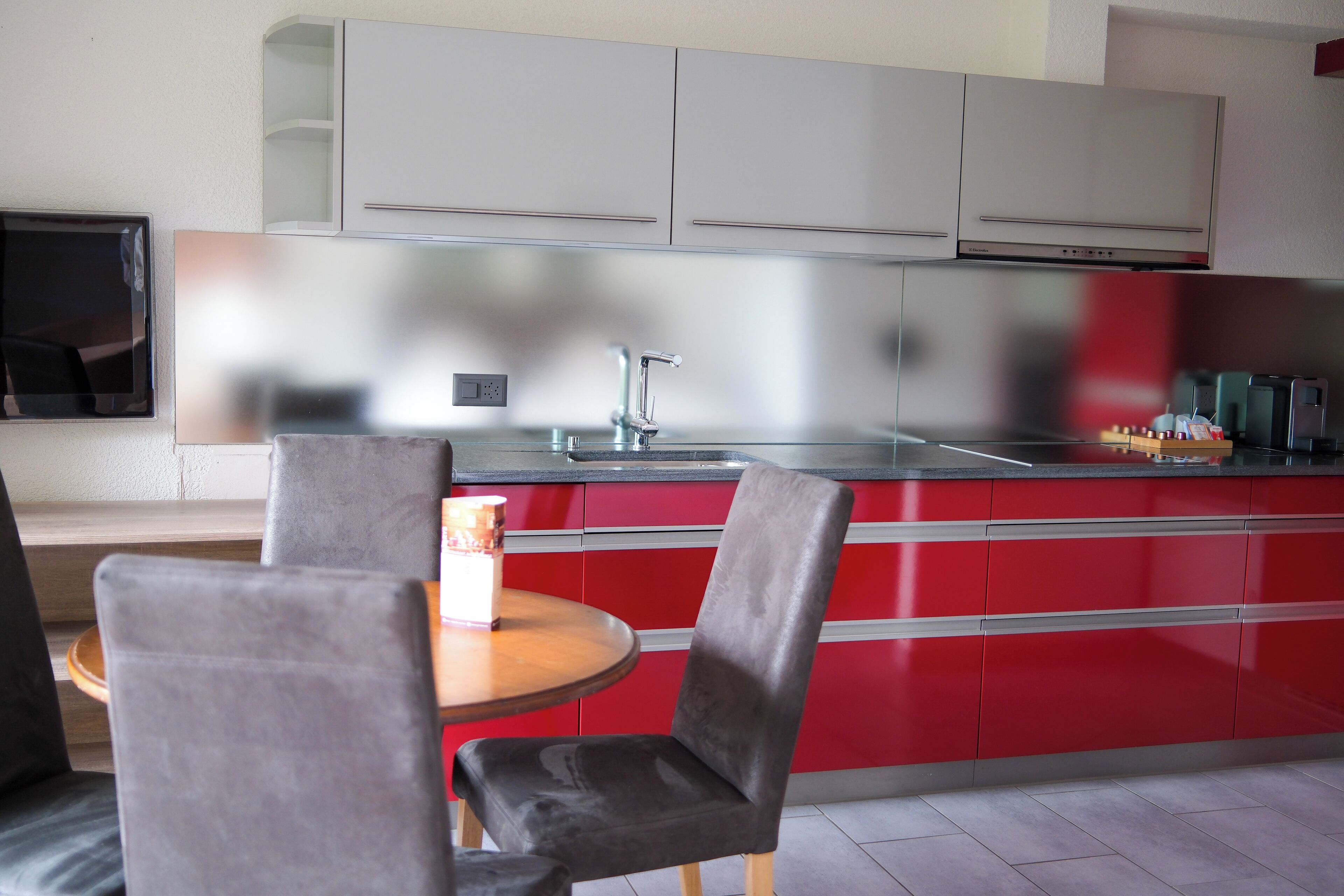 Cozinha moderna com armários vermelhos e brancos, uma pia de aço inoxidável e um espaço de jantar com mesa redonda e duas cadeiras.