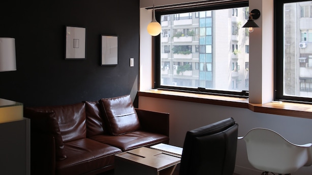 Quarto moderno com sofá de couro, uma cadeira preta, uma cadeira branca, uma mesinha e uma janela com vista para edifícios.
