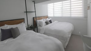 Quarto de hotel com duas camas, uma mesinha de cabeceira, uma cadeira e janela com persianas.