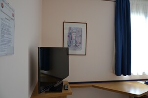 Quarto de hotel com televisão de tela plana sobre um suporte de madeira, um quadro na parede e uma janela com cortinas azuis.