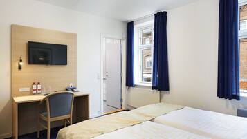 Quarto de hotel com cama, escrivaninha com cadeira, televisão presa na parede e janela com cortinas.