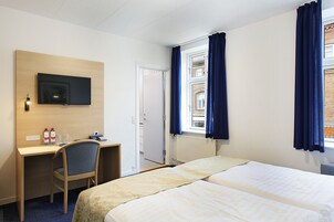 Quarto de hotel com cama, escrivaninha com cadeira, televisão presa na parede e janela com cortinas.