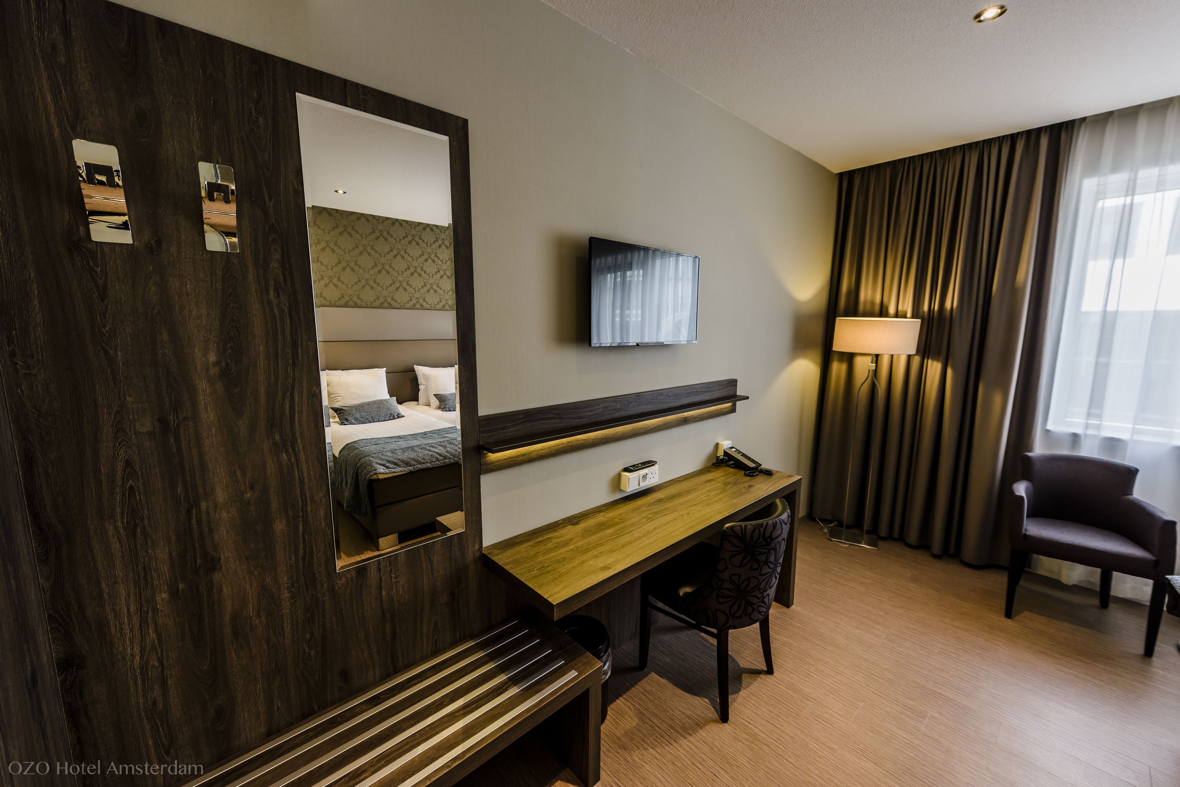 Quarto de hotel com cama, escrivaninha, cadeira, luminária e um espelho grande.