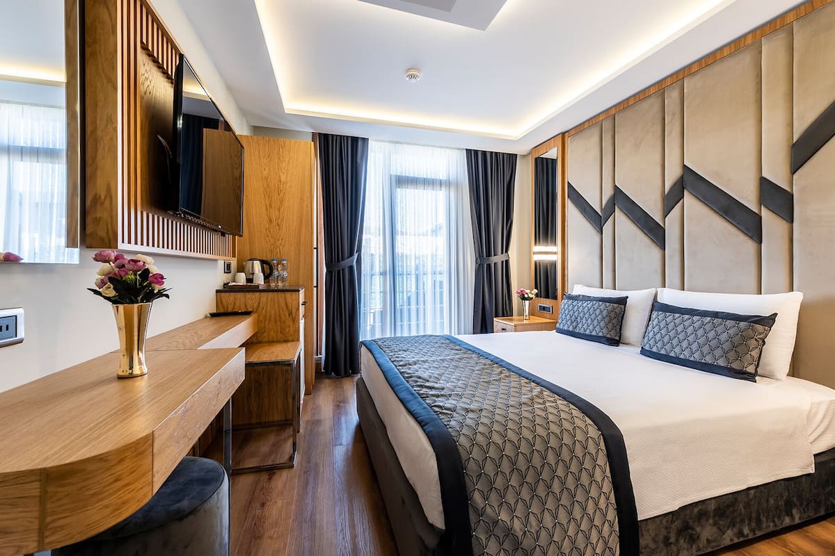 Quarto de hotel moderno com uma cama grande, uma escrivaninha de madeira, toucador e uma janela ampla com cortinas.