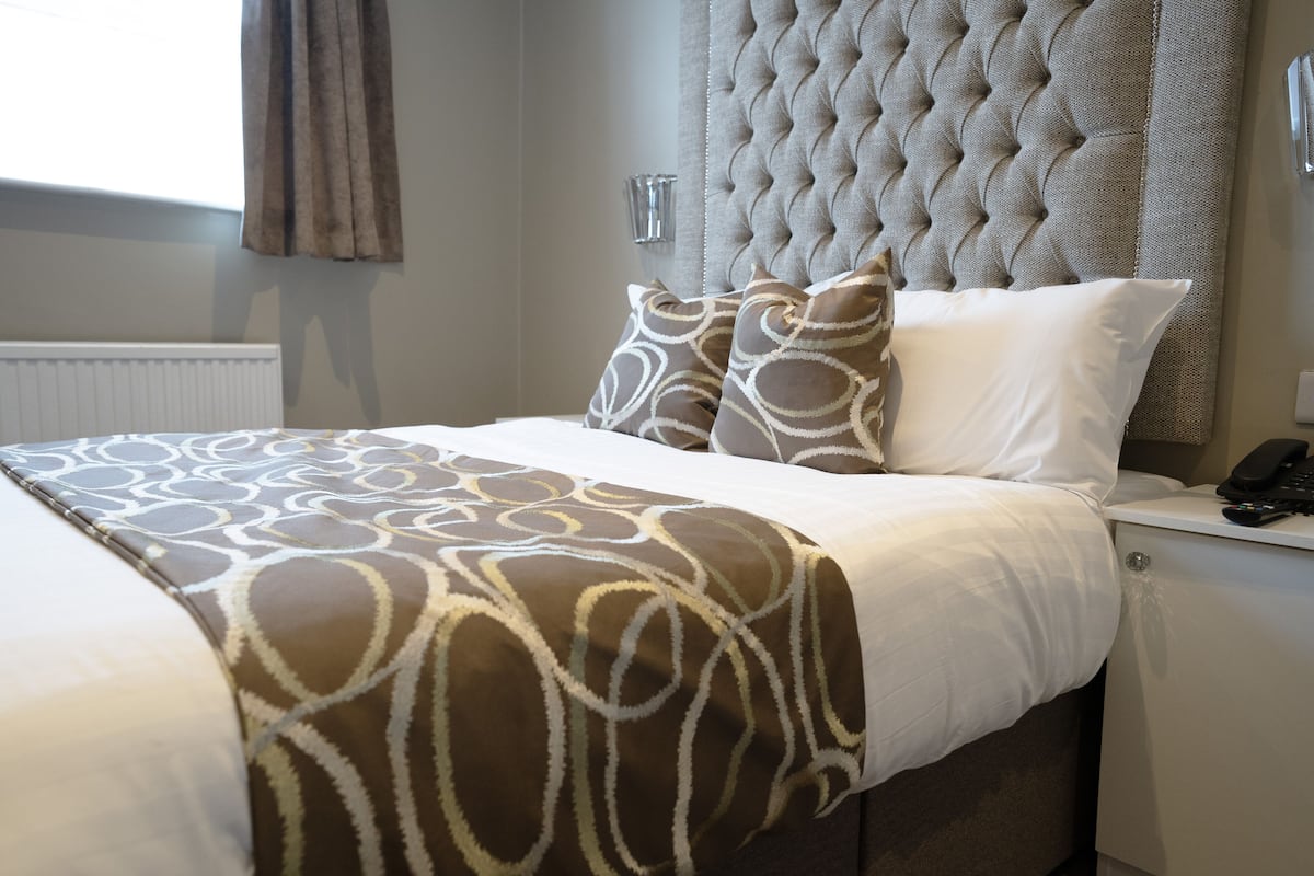 Cama bem arrumada com colcha estampada, cabeceira estofada e telefone ao lado da cama.