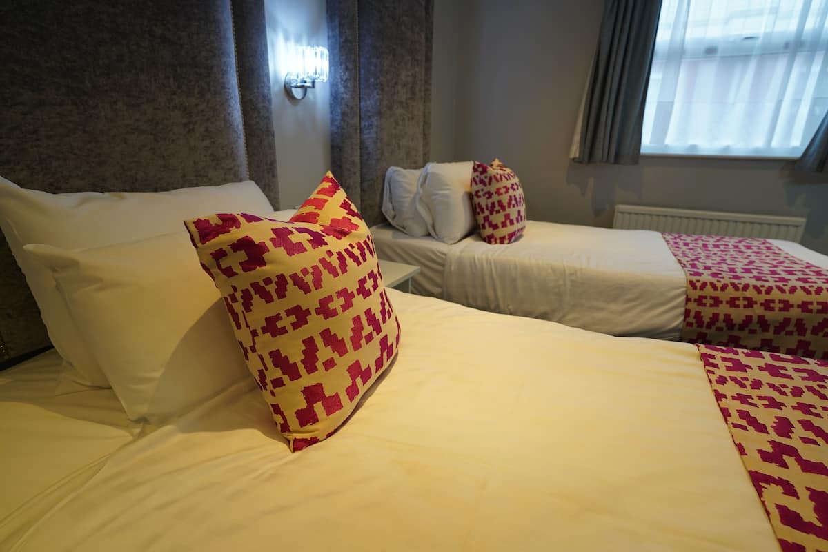 Quarto de hotel com duas camas, cada uma com roupa de cama branca e almofadas estampadas. Há uma janela com cortinas e um candeeiro fixo na parede.