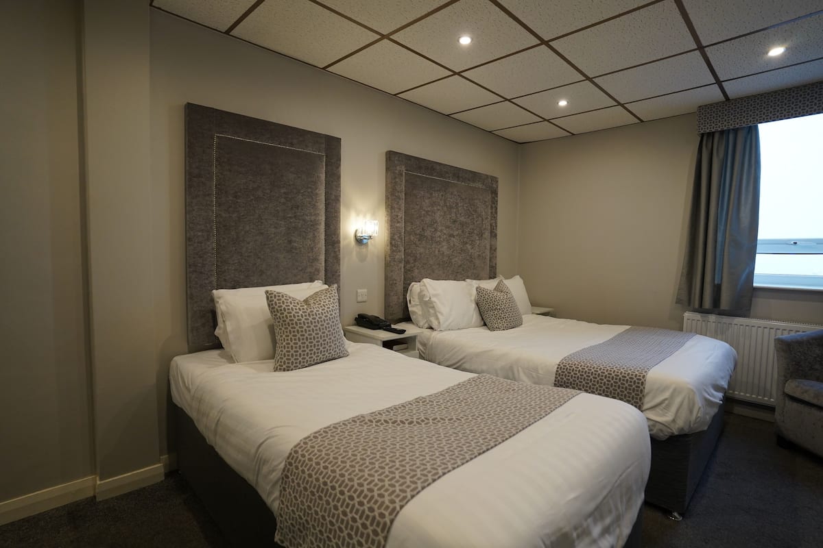 Quarto de hotel com duas camas, uma mesa de cabeceira, uma cadeira e uma janela com cortinas.
