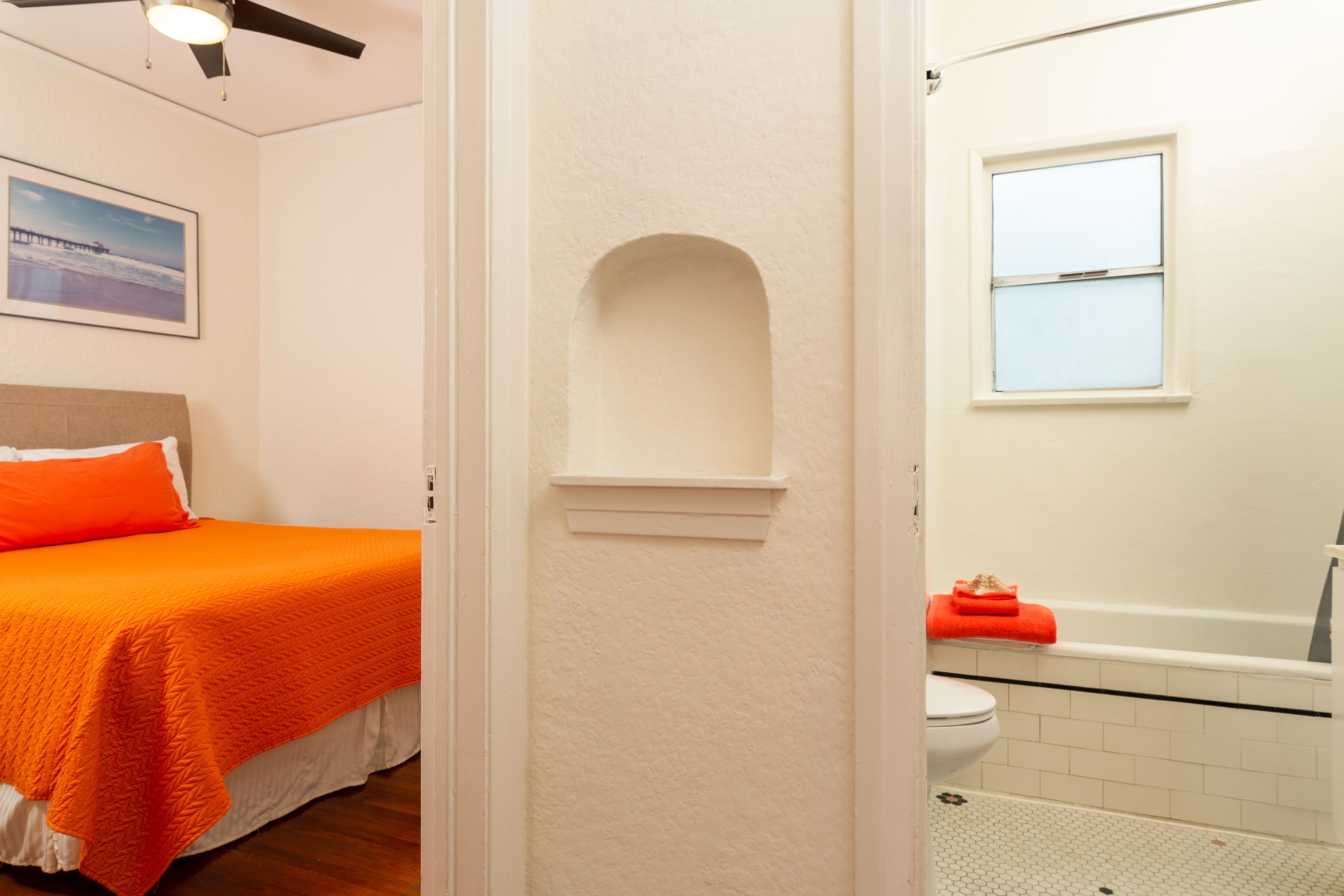 Quarto com colcha laranja, parede branca com prateleira embutida e banheiro com banheira e janela.