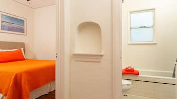 Quarto com colcha laranja, parede branca com prateleira embutida e banheiro com banheira e janela.