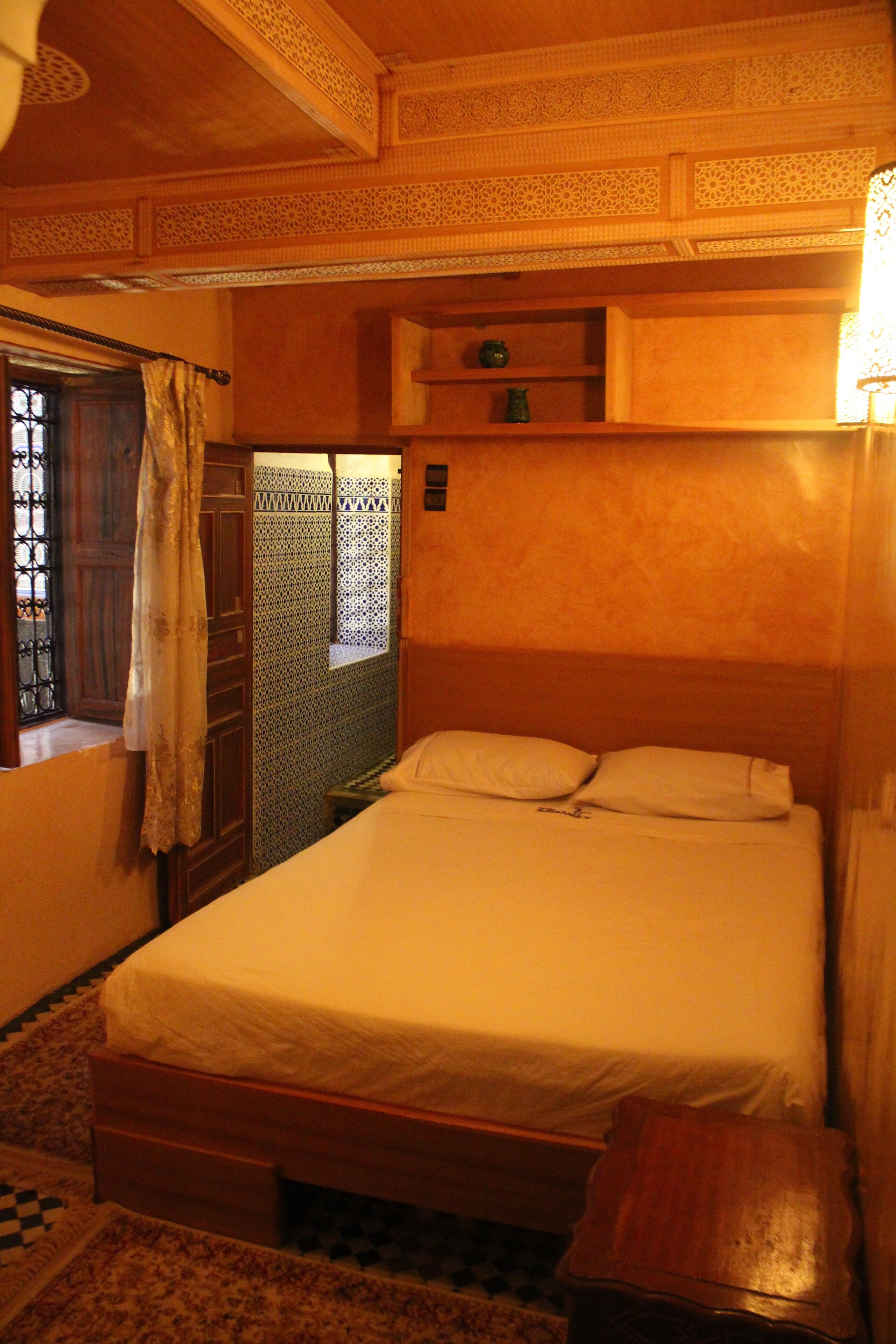 Um quarto com uma cama, uma pequena mesa de cabeceira de madeira, uma janela com cortina e uma estante com um objeto decorativo.