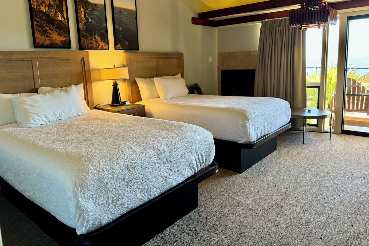 Quarto de hotel com duas camas, lareira, varanda com vista e obras de arte na parede.