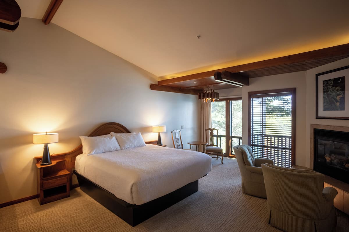 Quarto de hotel com uma cama grande, duas mesas de cabeceira com luminárias, uma mesinha redonda, duas poltronas, uma lareira e uma janela com persianas.