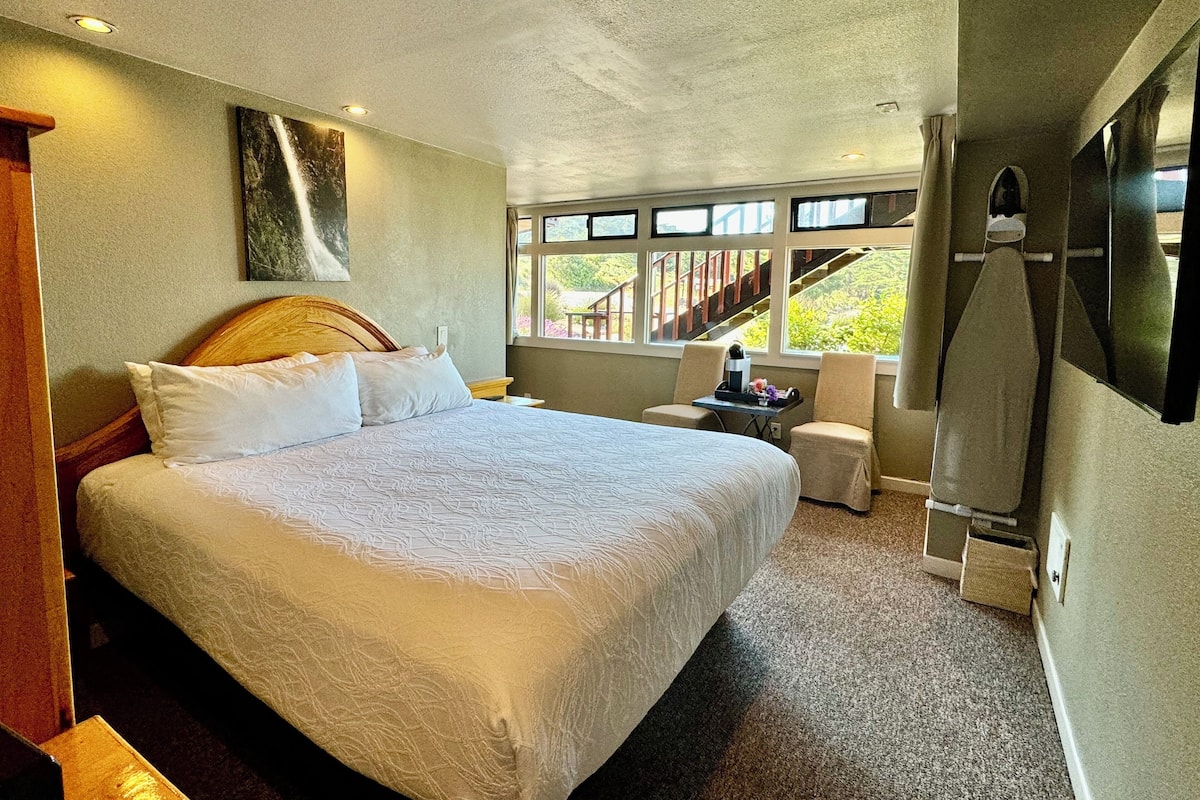 Um quarto espaçoso com uma cama grande, uma mesinha com cadeira e vista para uma escadaria que dá acesso a áreas externas.