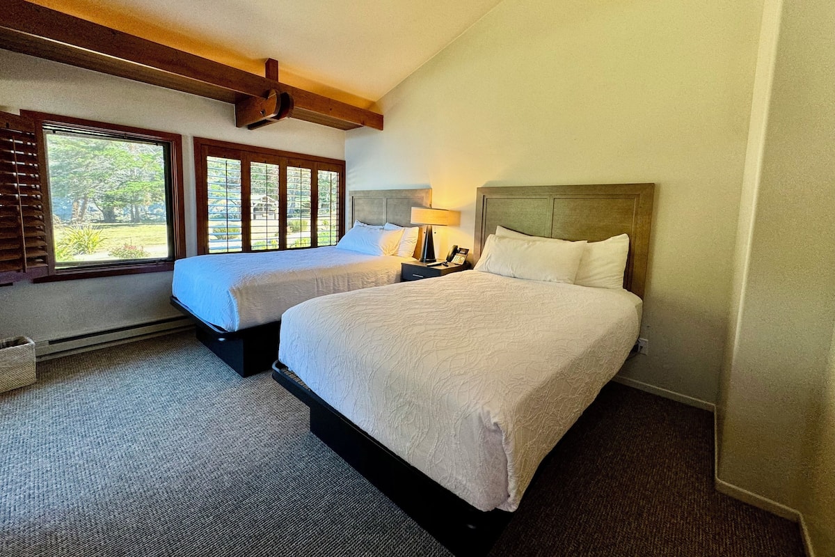 Quarto de hotel com duas camas, ventoinha no teto, janela com persianas e mesa de cabeceira com abajur.