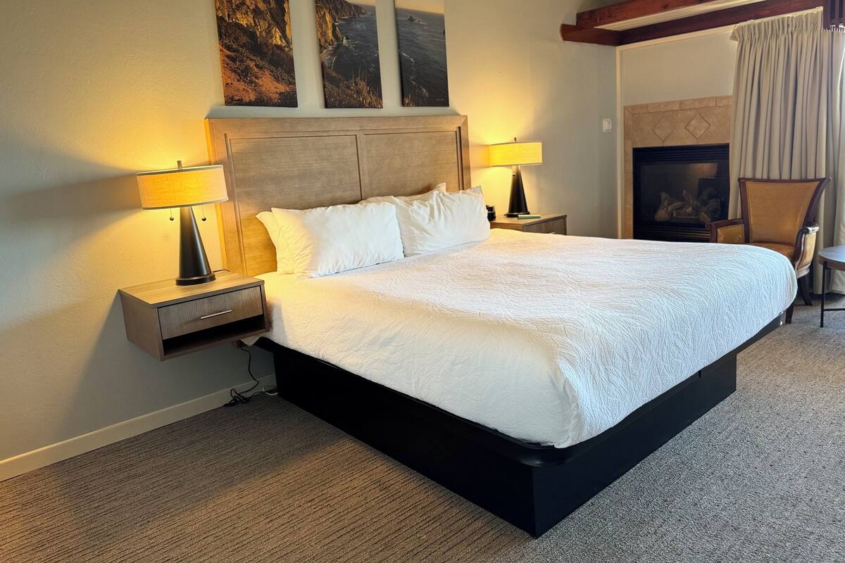 Quarto de hotel com uma cama grande, duas mesas de cabeceira com luminárias, uma lareira e três quadros na parede.