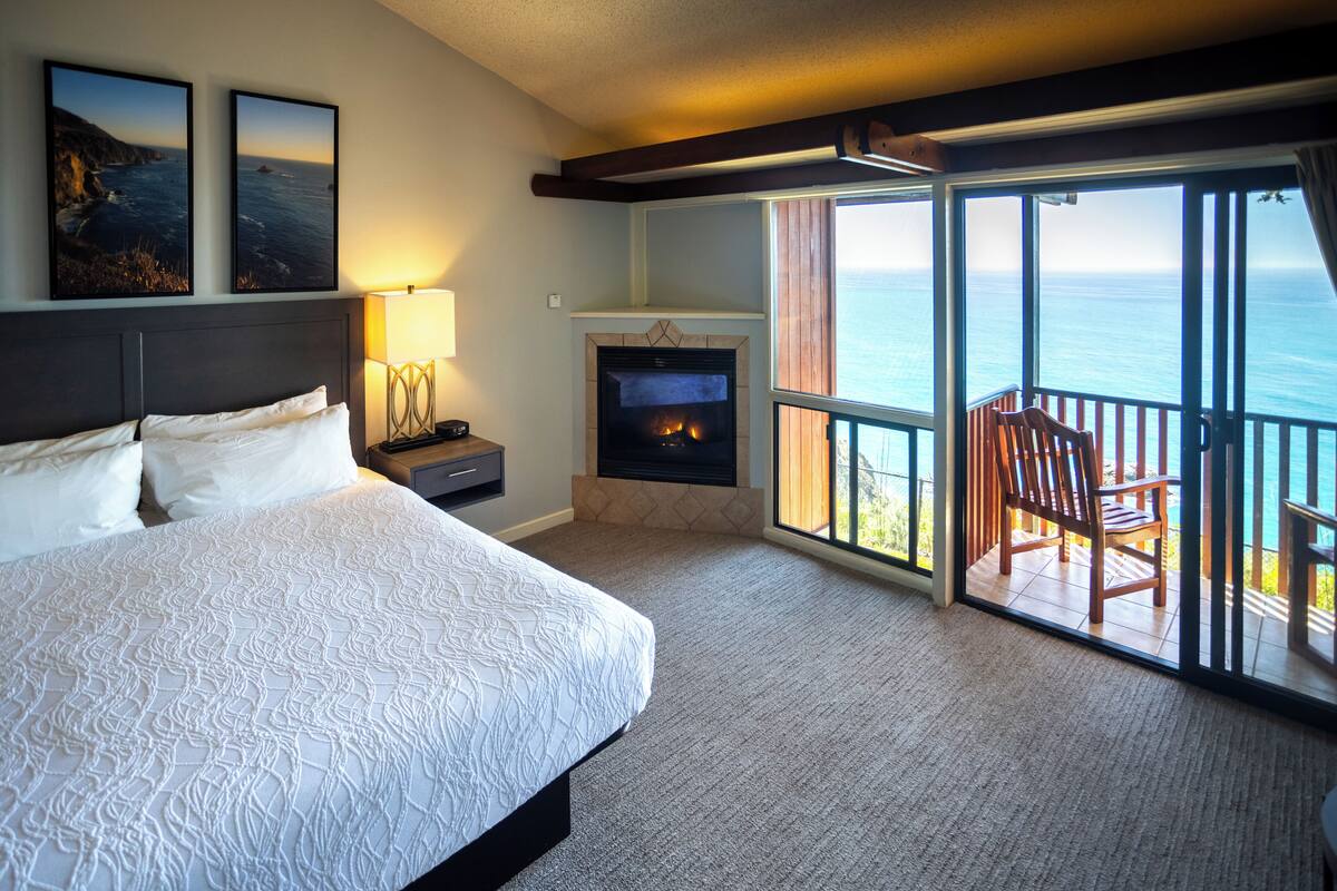 Quarto de hotel com uma cama grande, uma lareira, uma varanda com vista para o oceano e duas fotografias emolduradas na parede.