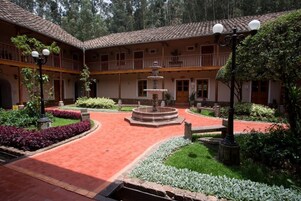 Pátio com uma fonte central, ladeado por um caminho de tijolos vermelhos e vegetação exuberante.