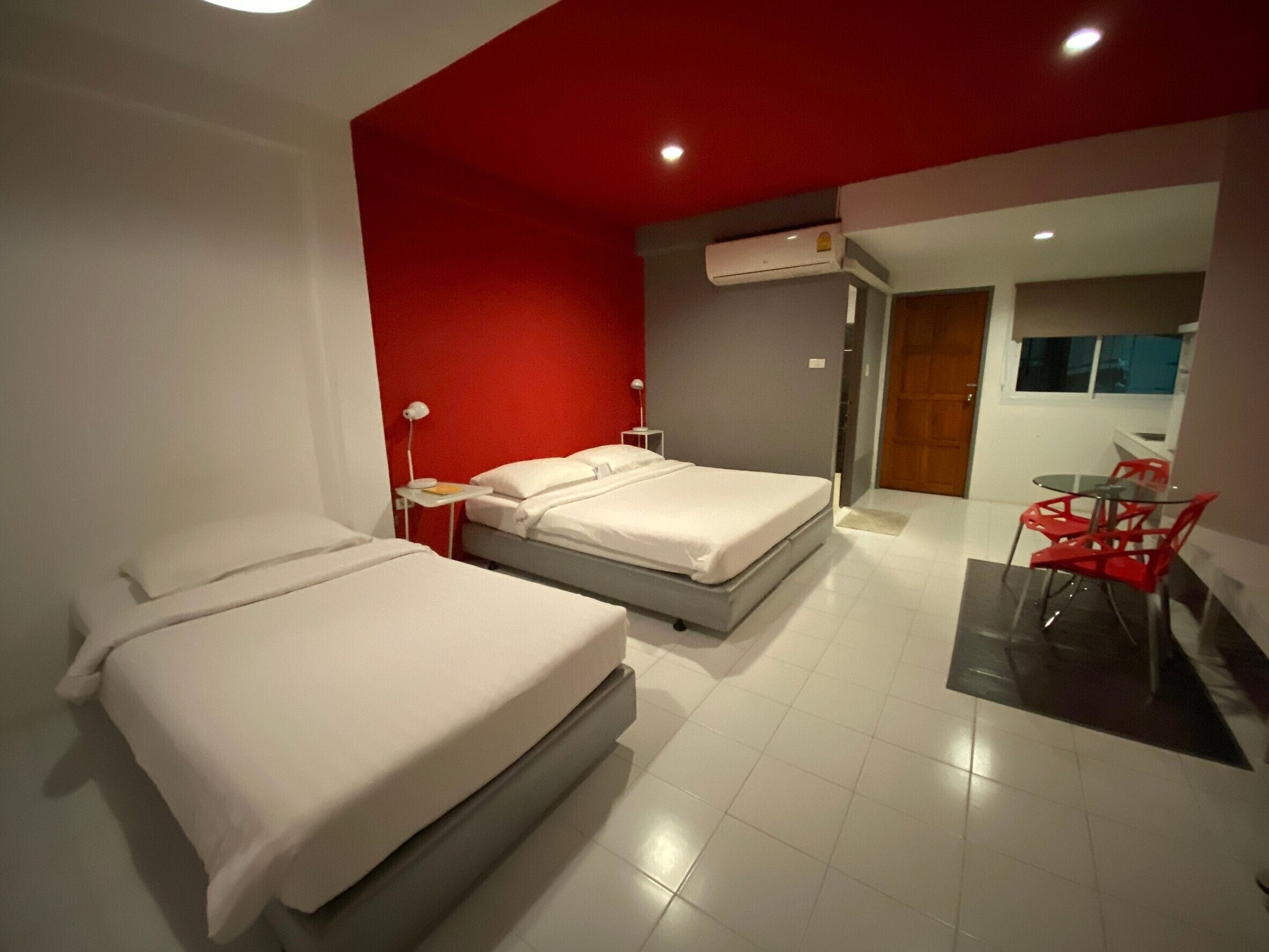 Quarto de hotel com duas camas, área de jantar com cadeiras vermelhas, parede de destaque vermelha e uma janela.